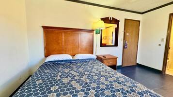 Quarto de hotel com uma cama grande, cabeceira de madeira, mesa de cabeceira com abajur e uma porta.