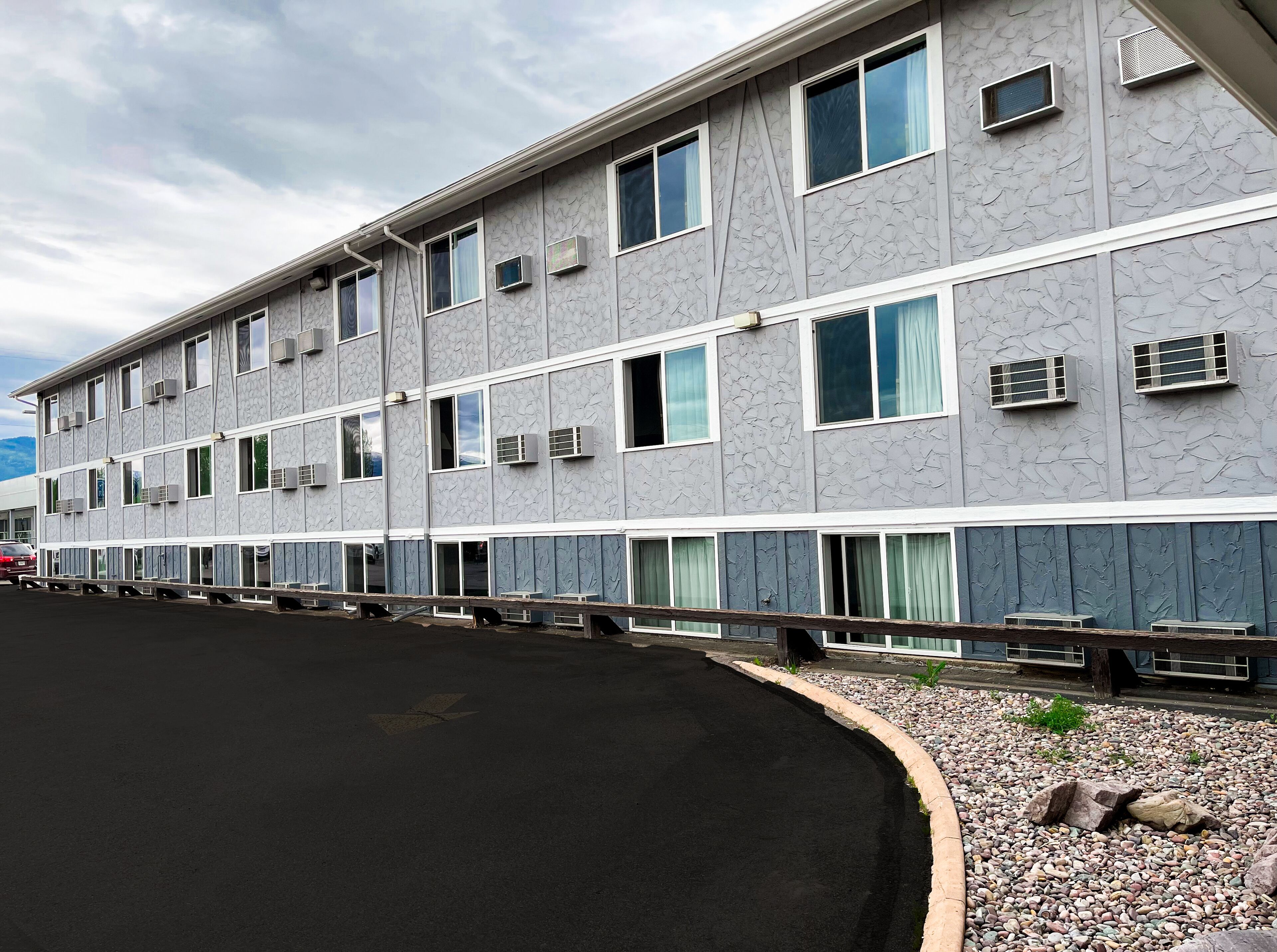Um hotel com vários andares, fachada moderna, unidades de ar condicionado e área de estacionamento com caminho de cascalho.