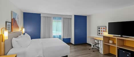 Quarto de hotel com cama, escrivaninha, televisão e um pequeno frigorífico.