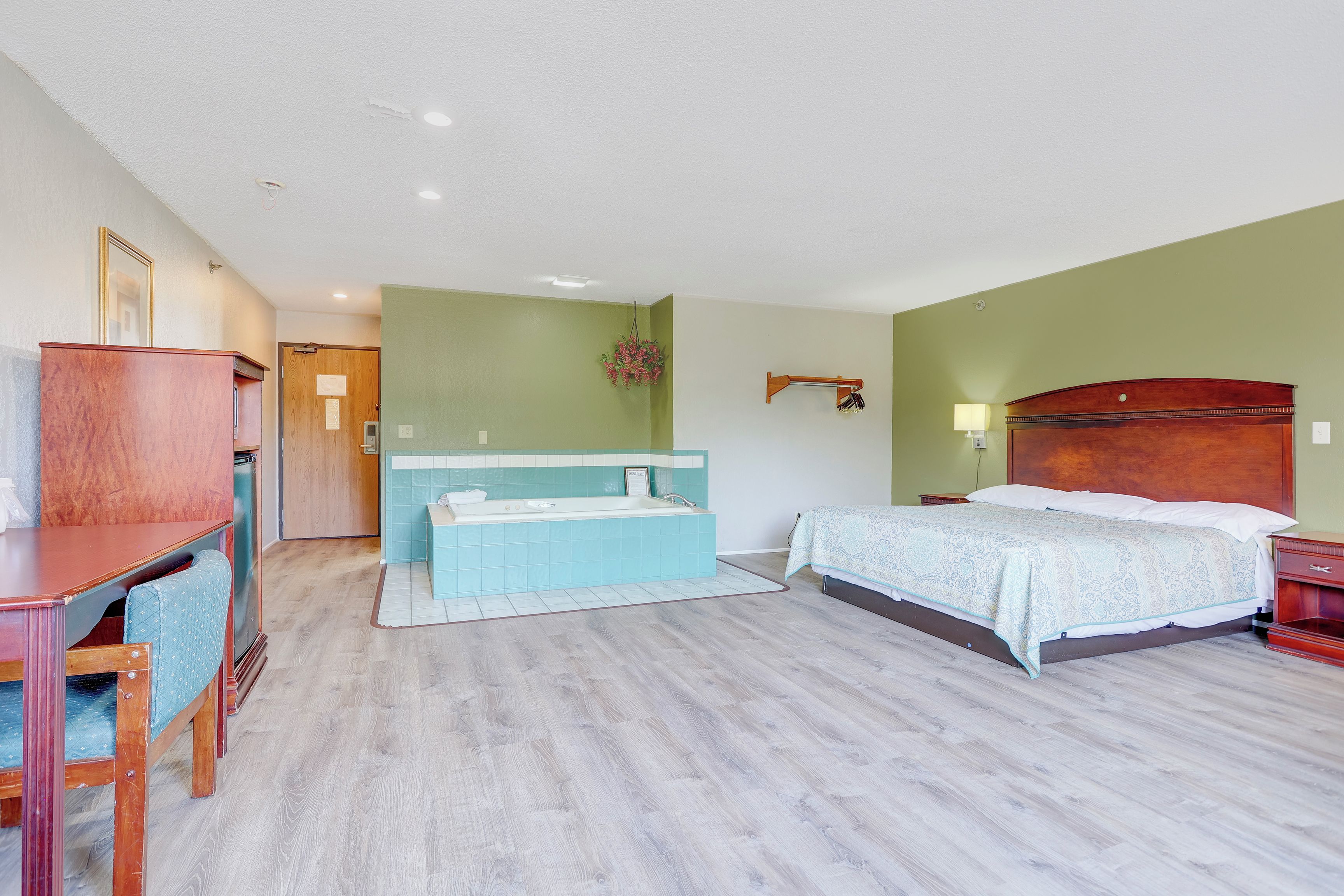 Quarto de hotel com uma secretária de madeira, uma cama grande, banheiro com banheira e uma parede verde.