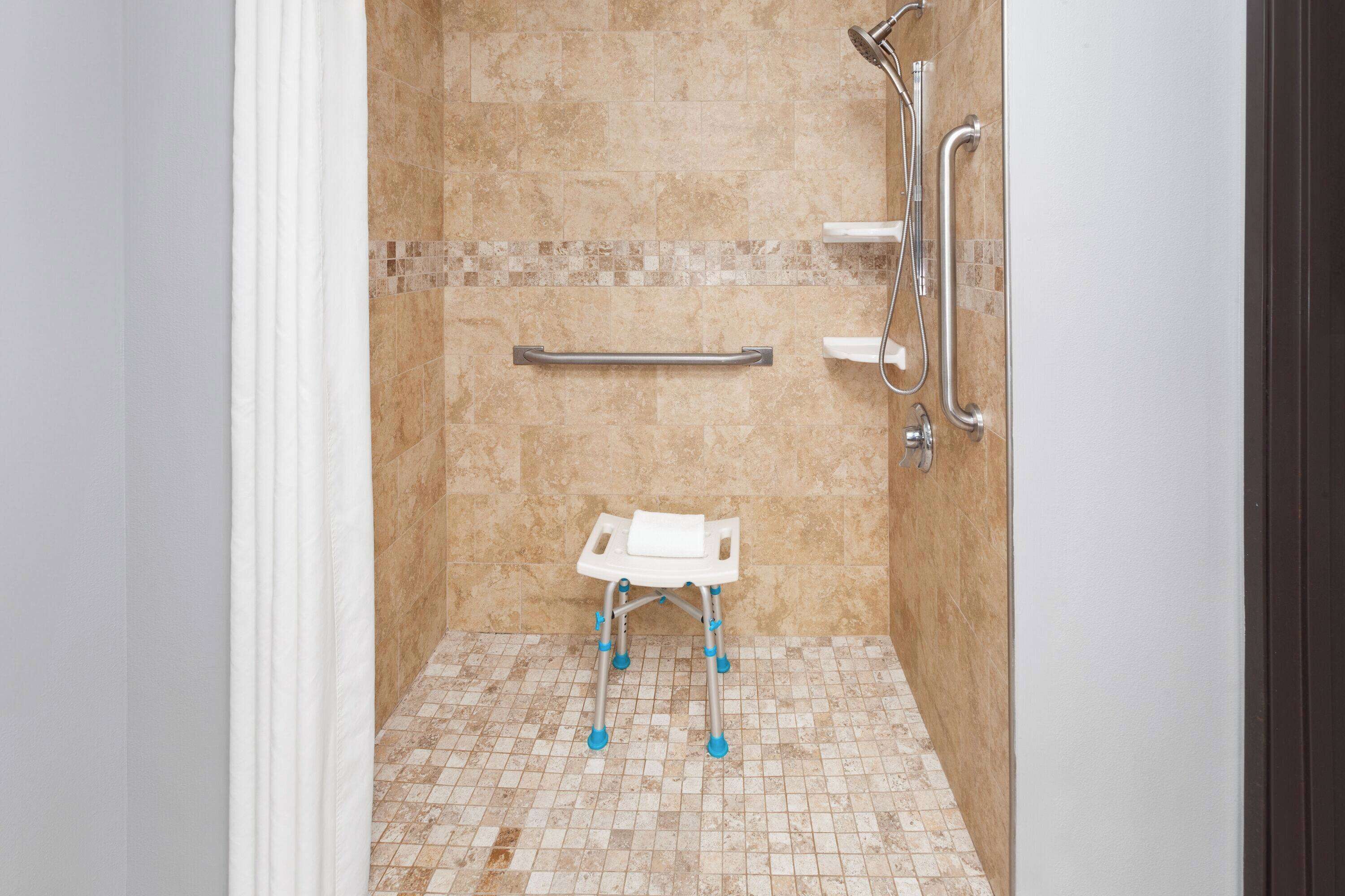 Área de chuveiro com barra de apoio, assento branco e piso revestido de azulejos.