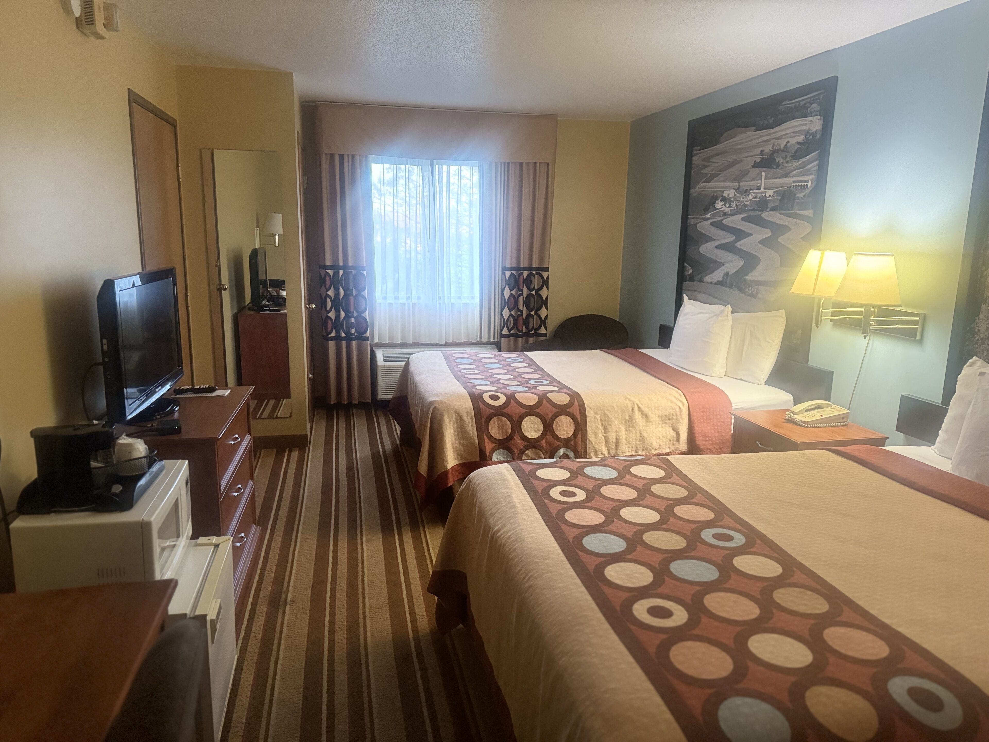 Quarto de hotel com duas camas, televisão, escrivaninha com computador e janela com cortinas.