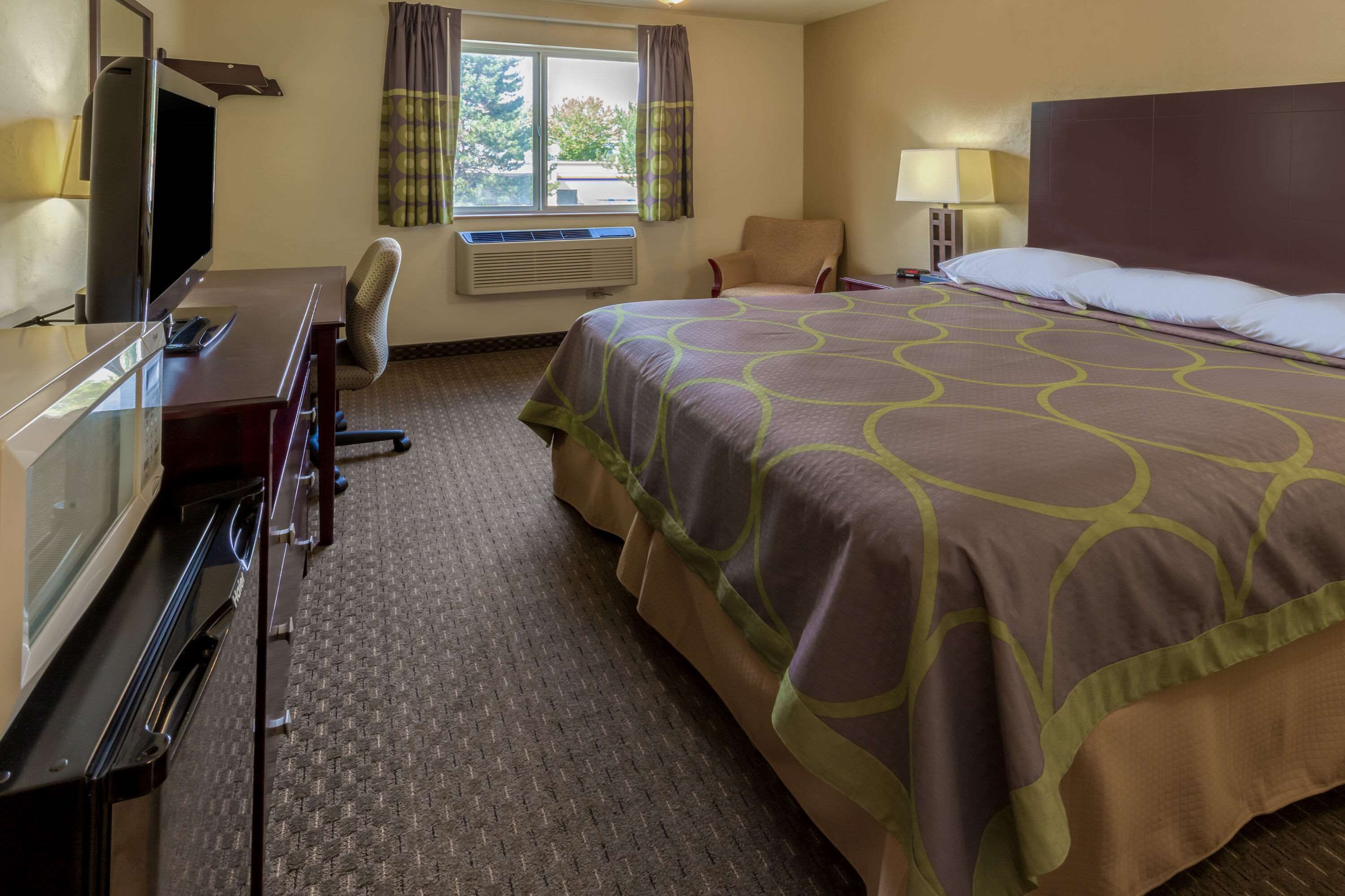 Quarto de hotel com cama, escrivaninha com televisão, cadeira e janela com cortinas.