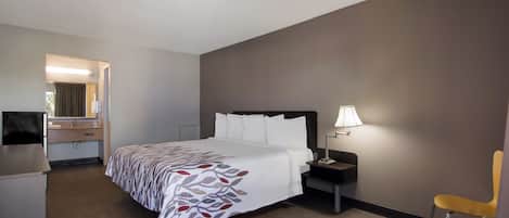 Quarto de hotel com cama, escrivaninha, cadeira, mesa de cabeceira com abajur e espelho.