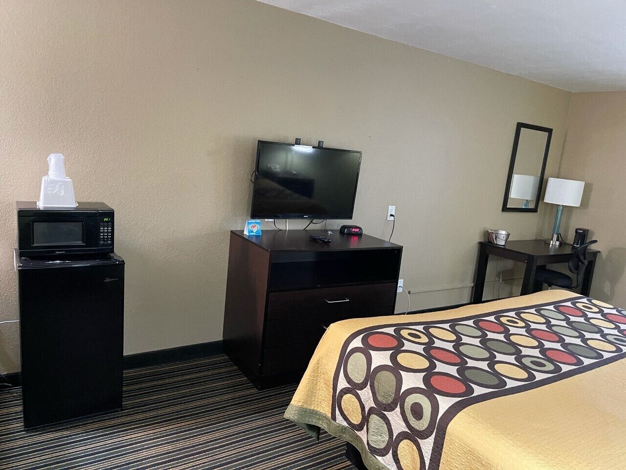 Quarto de hotel com cama, televisão em uma cômoda, micro-ondas e escrivaninha com abajur.