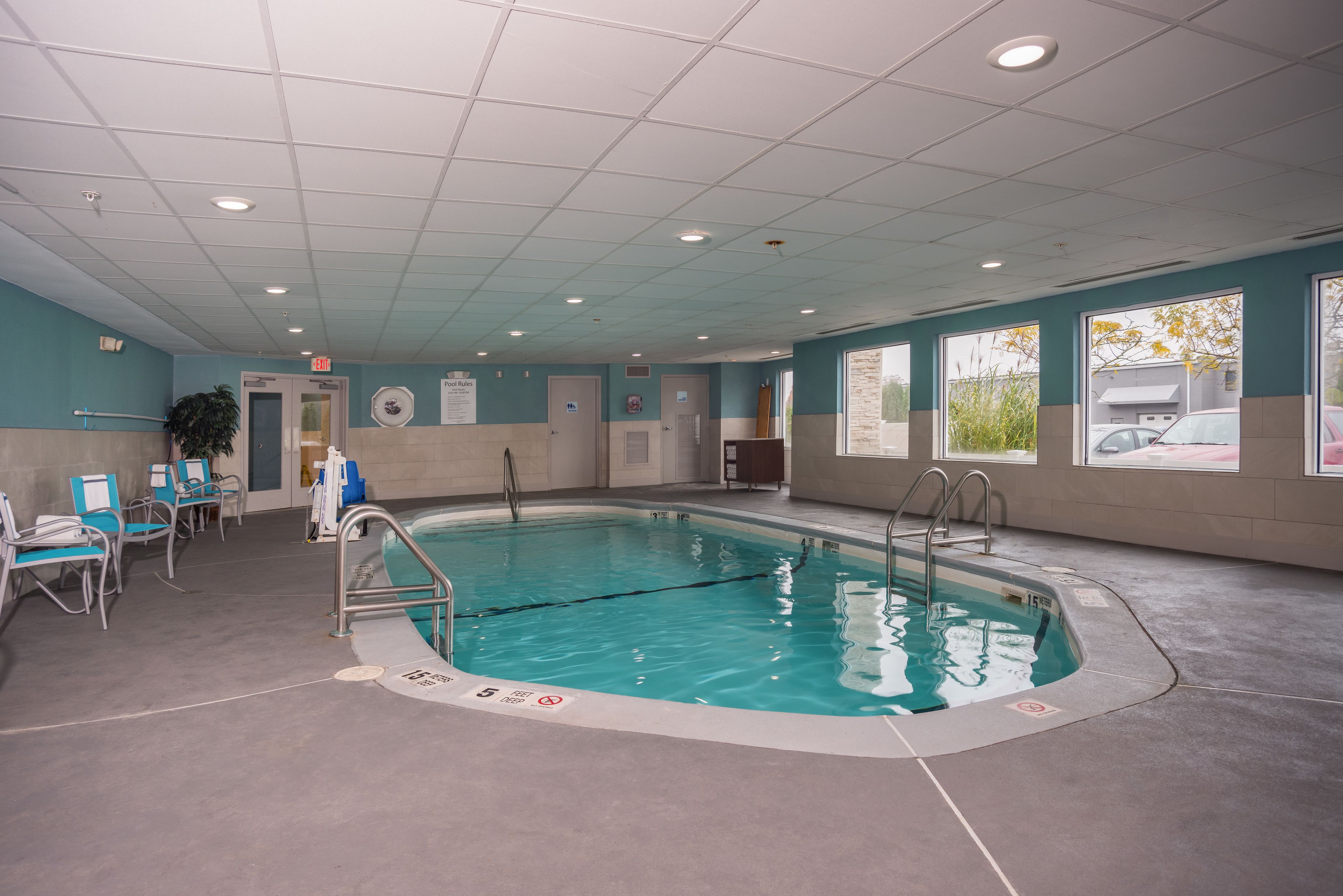 Uma piscina interna com água cristalina, ladeada por piso e paredes azulejadas. Há cadeiras e um banco ao redor da piscina, e uma porta que dá para outro cômodo.