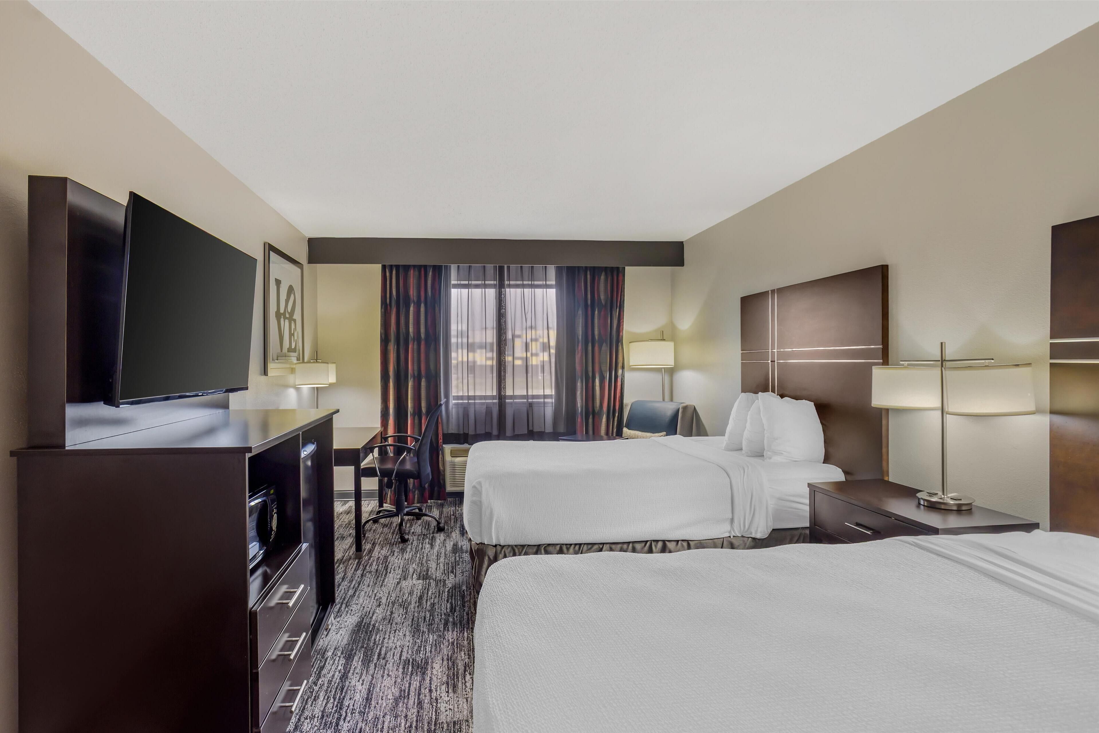 Quarto de hotel com duas camas, uma escrivaninha com televisão de tela plana, uma cadeira e uma luminária de mesa.