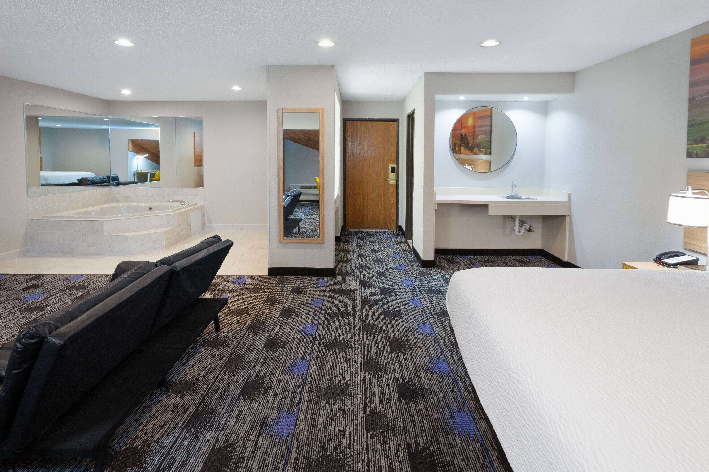 Quarto de hotel com uma cama grande, área de estar separada com uma cadeira, banheira, espelho amplo, pia e porta que dá para outro cômodo.