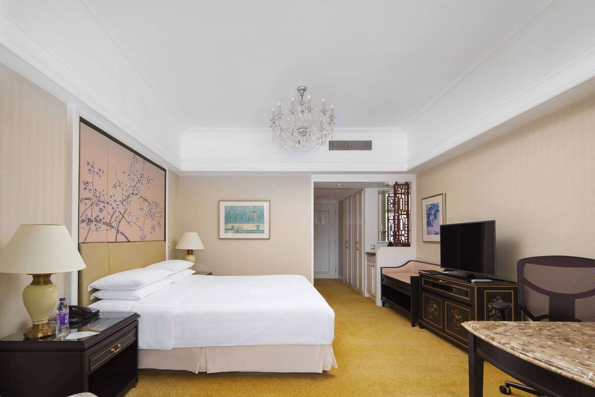 Quarto de hotel com cama, mesas de cabeceira, escrivaninha com cadeira, televisão e obras de arte nas paredes.