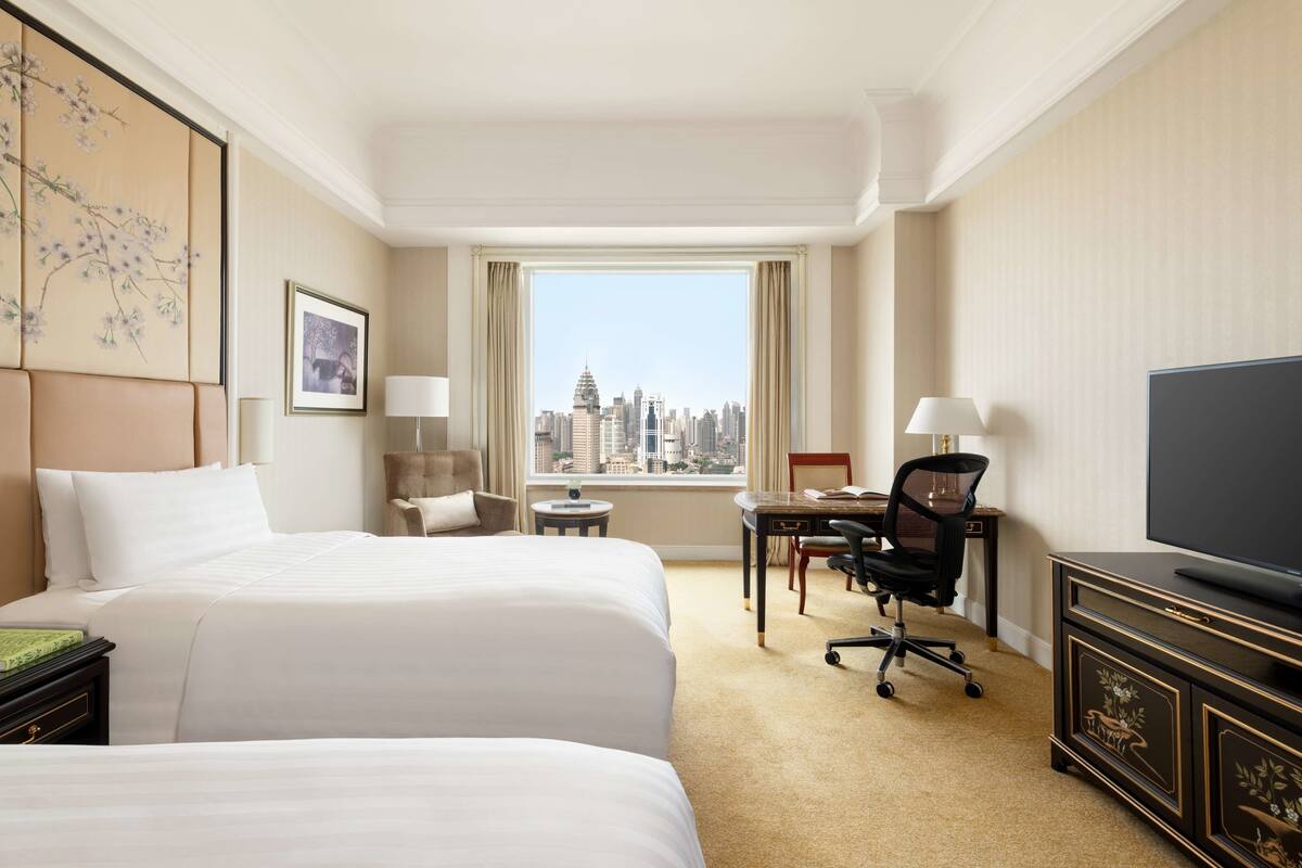 Quarto de hotel com duas camas, uma escrivaninha, uma cadeira, televisão e vista para o horizonte da cidade.