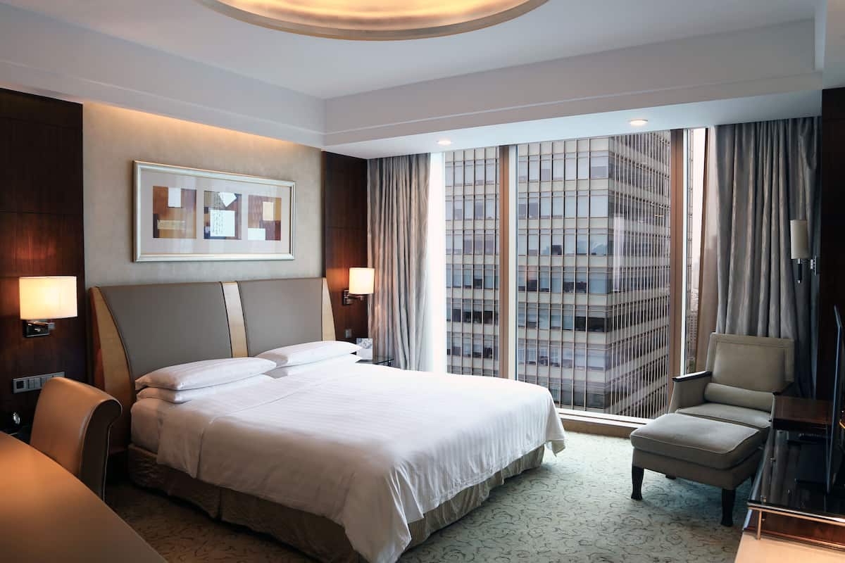 Quarto de hotel com uma cama grande, uma cadeira, uma escrivaninha e vista para a paisagem urbana através de grandes janelas.
