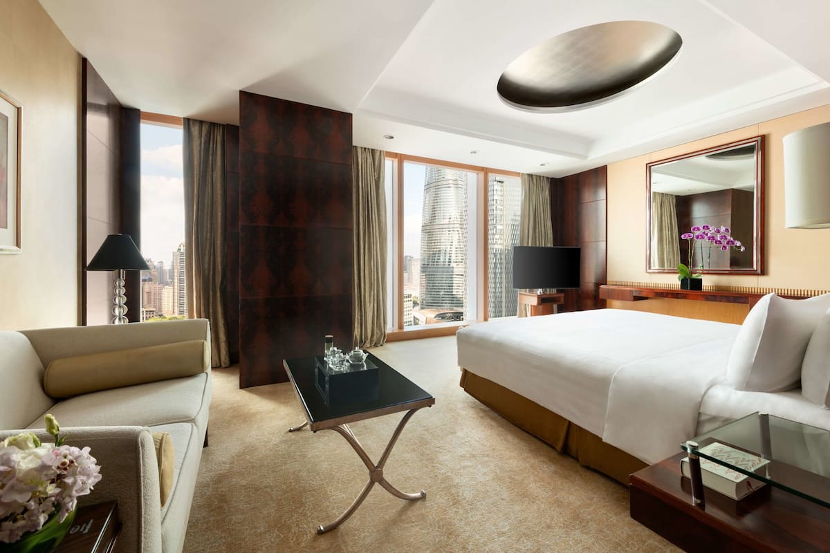 Quarto de hotel com uma cama grande, um sofá, uma mesa de centro de vidro e vista para edifícios altos.