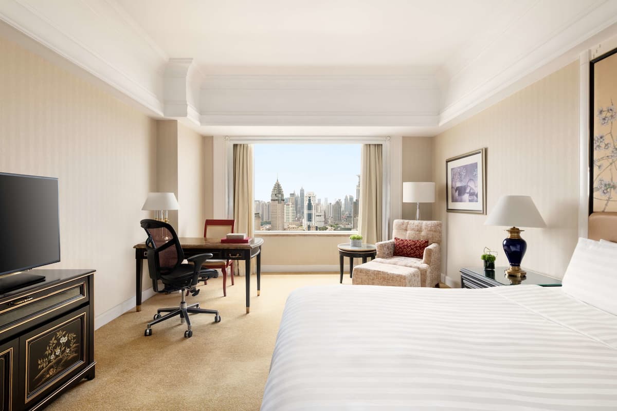 Quarto de hotel com uma cama grande, uma escrivaninha com cadeira, televisão e vista para o horizonte da cidade.