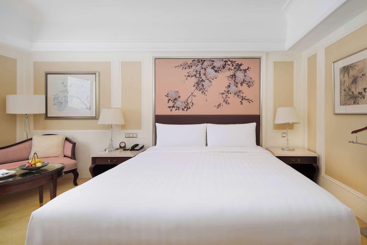 Quarto de hotel com uma cama grande, duas mesas de cabeceira com luminárias, uma cadeira e uma mesinha com uma cesta de frutas.