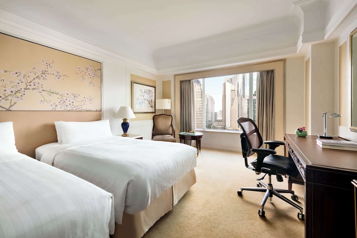 Quarto de hotel com duas camas, uma escrivaninha, uma cadeira, um abajur, um quadro e uma janela com vista para a cidade.