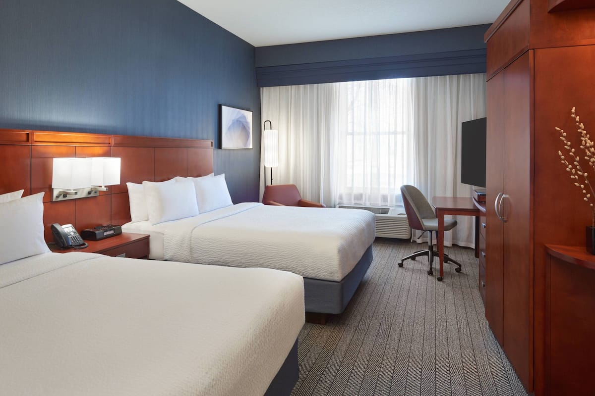 Quarto de hotel com duas camas, uma escrivaninha, uma cadeira, uma televisão e um telefone.