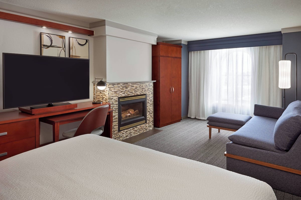 Quarto de hotel com cama, escrivaninha com televisão de tela plana, lareira, sofá e uma janela grande com cortinas.
