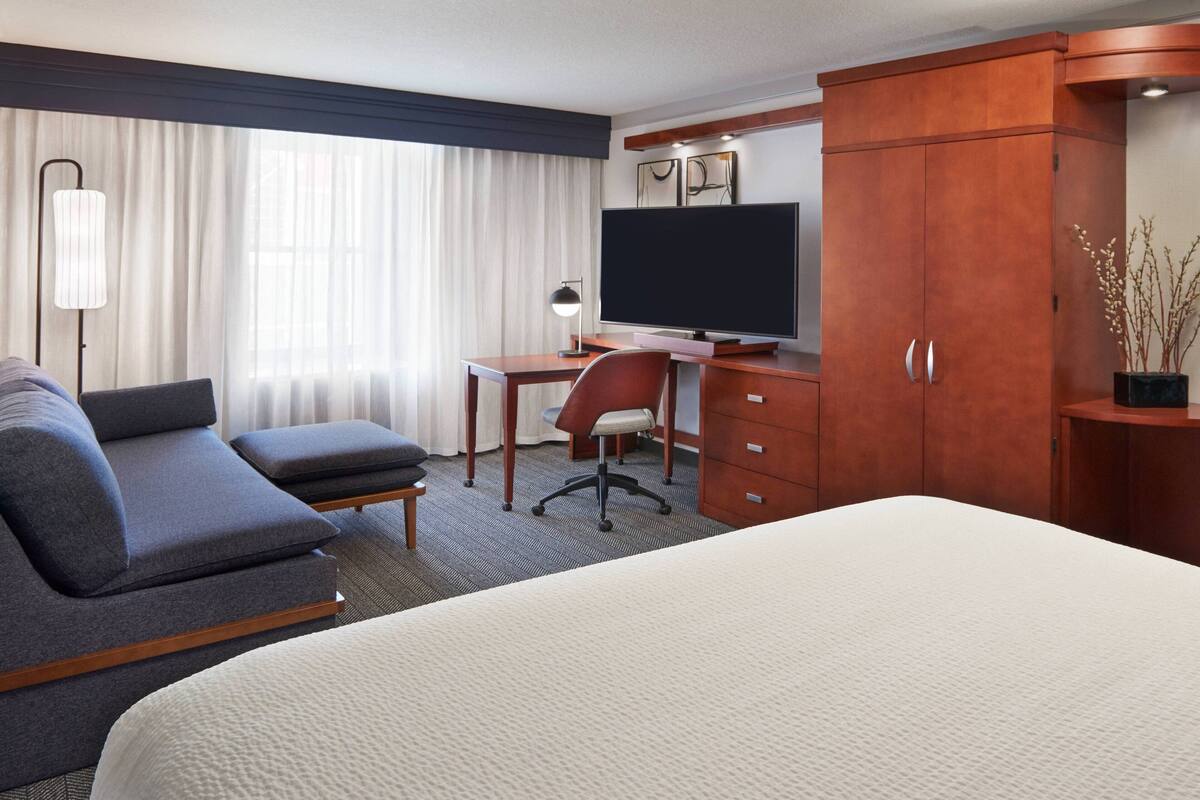 Quarto de hotel com uma cama grande, uma escrivaninha com televisão de tela plana, um sofá, uma cadeira e um guarda-roupa.