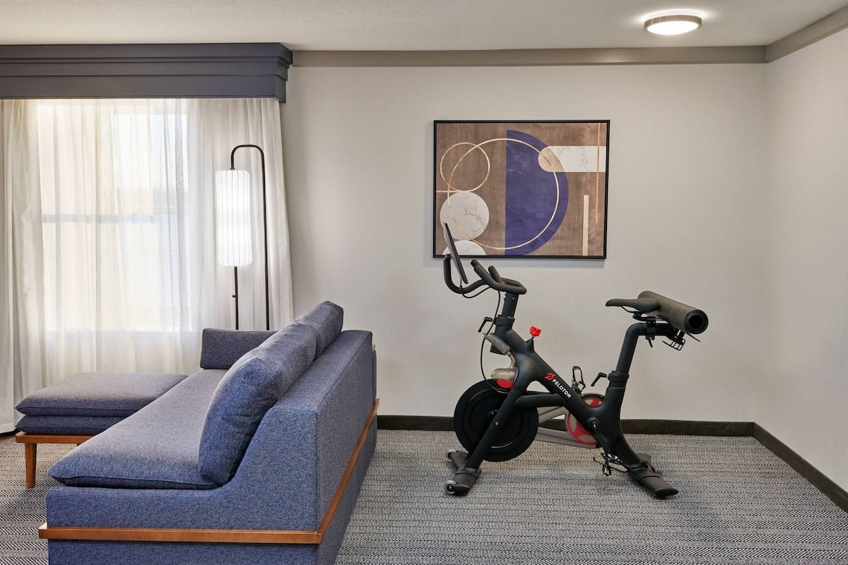 Quarto de hotel com sofá cinzento, luminária de chão e bicicleta ergométrica.
