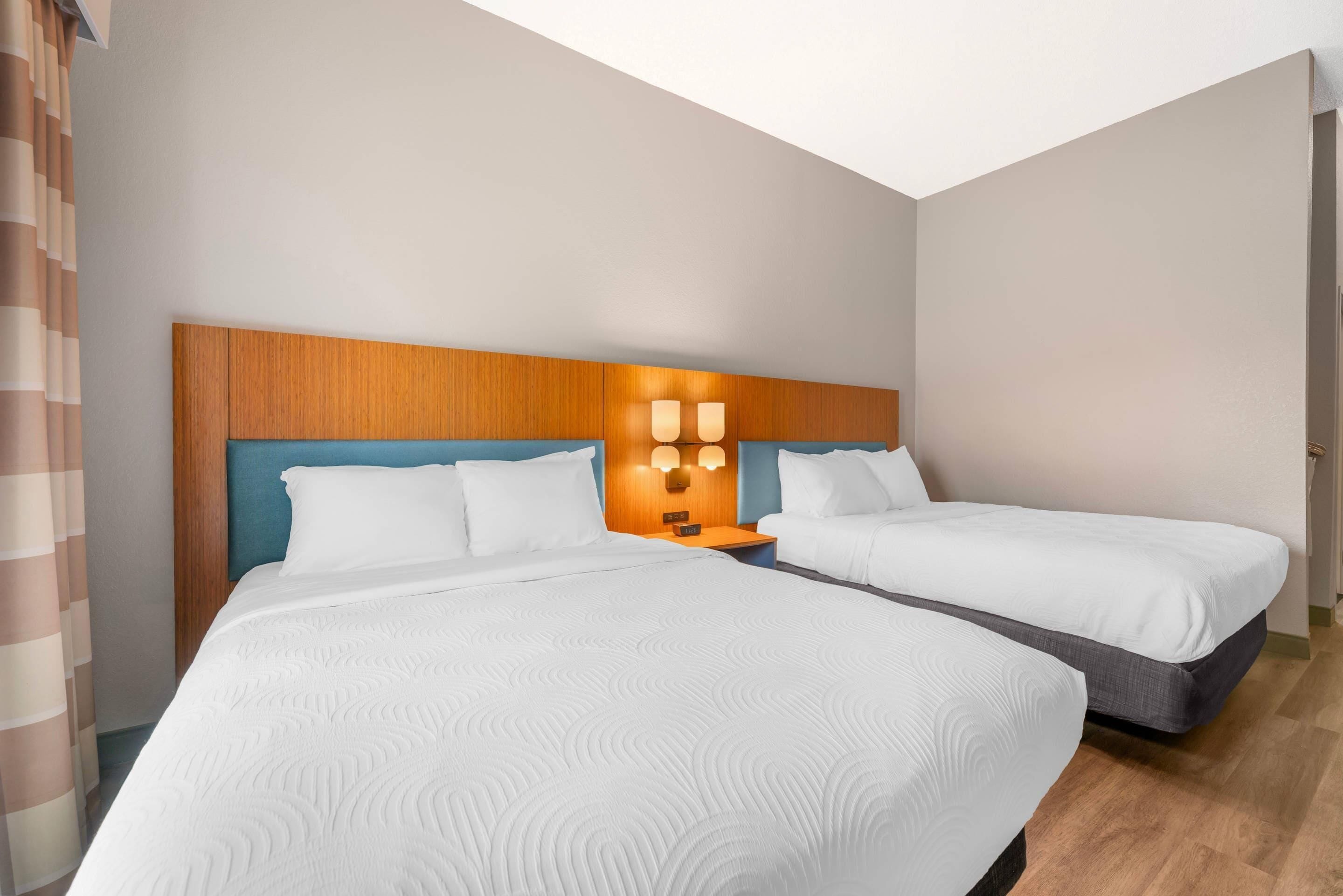 Quarto de hotel com duas camas, cabeceira de madeira, luminárias fixadas na parede e piso de madeira.