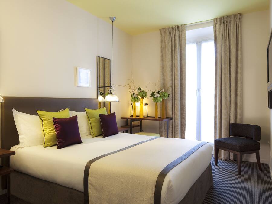 Quarto de hotel com uma cama grande, mesas de cabeceira, escrivaninha com luminária, uma cadeira e televisão na parede.