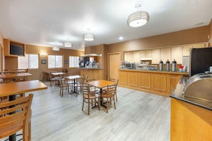 Uma ampla sala de jantar com mesas e cadeiras de madeira, uma cozinha com armários de madeira e uma geladeira grande.