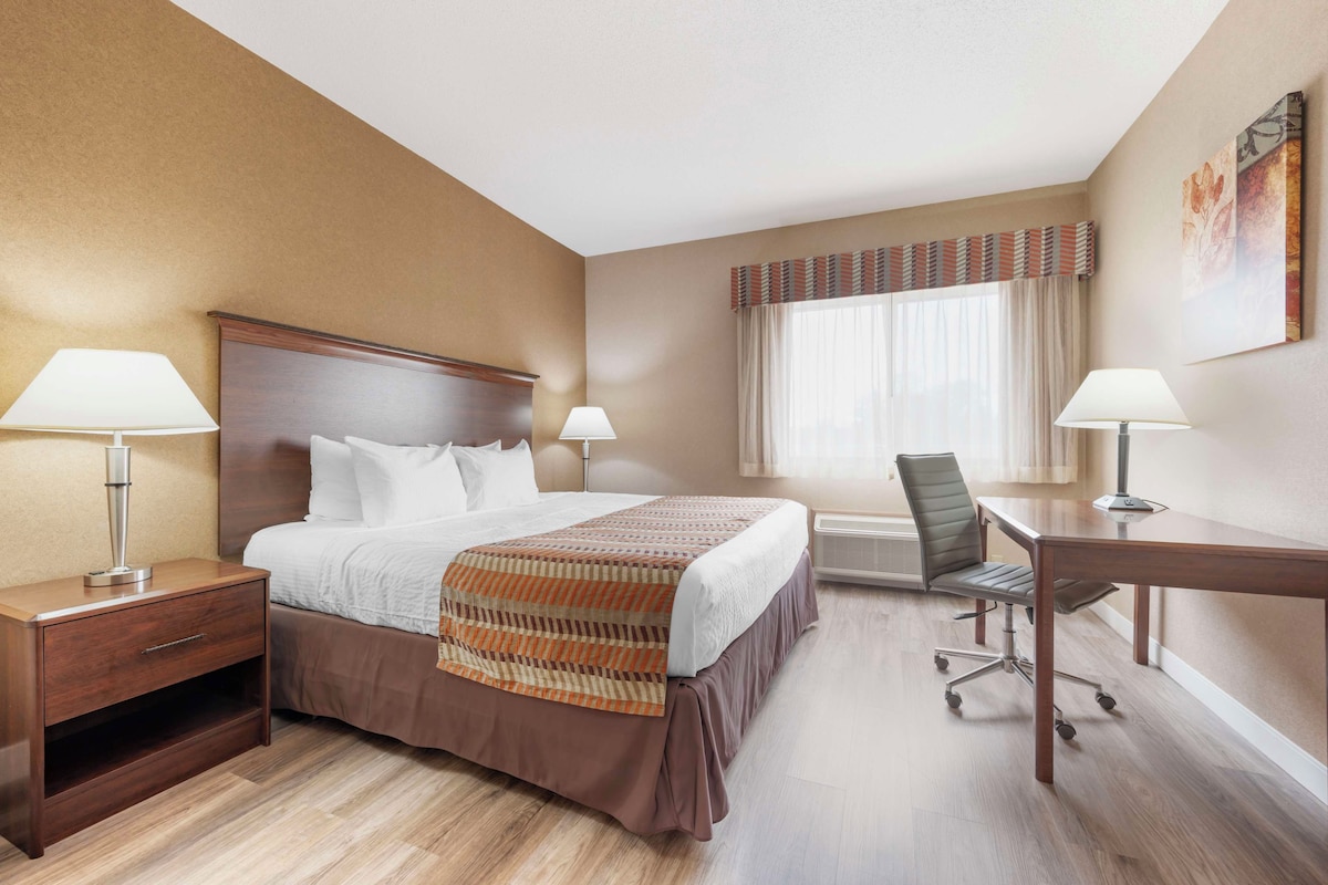 Quarto de hotel com uma cama grande, duas mesas de cabeceira com abajures, uma escrivaninha com cadeira e uma janela com cortinas.