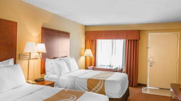 Quarto de hotel com duas camas, uma mesa de cabeceira, escrivaninha, luminária, janela com cortinas e porta.