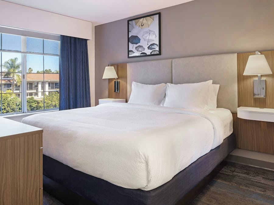 Quarto de hotel com uma cama grande, duas mesas de cabeceira com luminárias, vista para edifícios pela janela e uma moldura com imagem na parede.