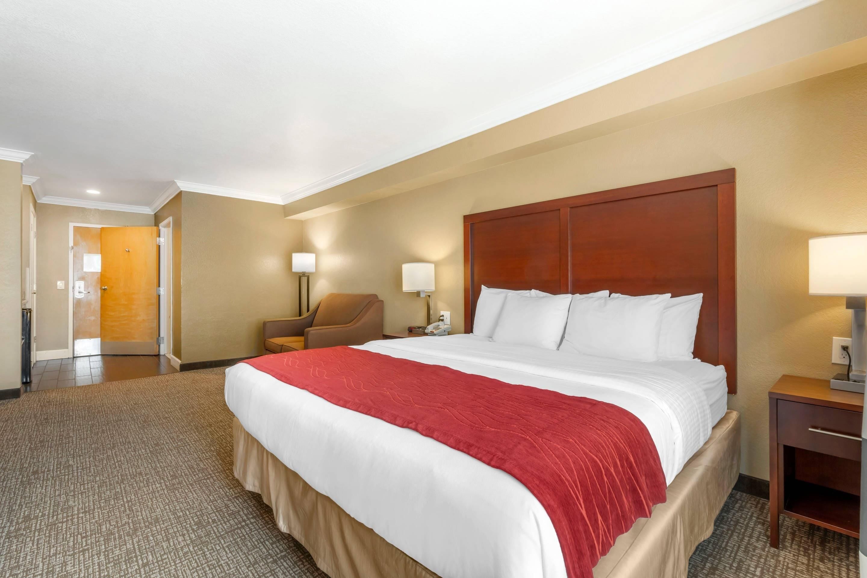 Quarto de hotel com uma cama grande, cabeceira de madeira, mesa de cabeceira com abajur e uma cadeira.