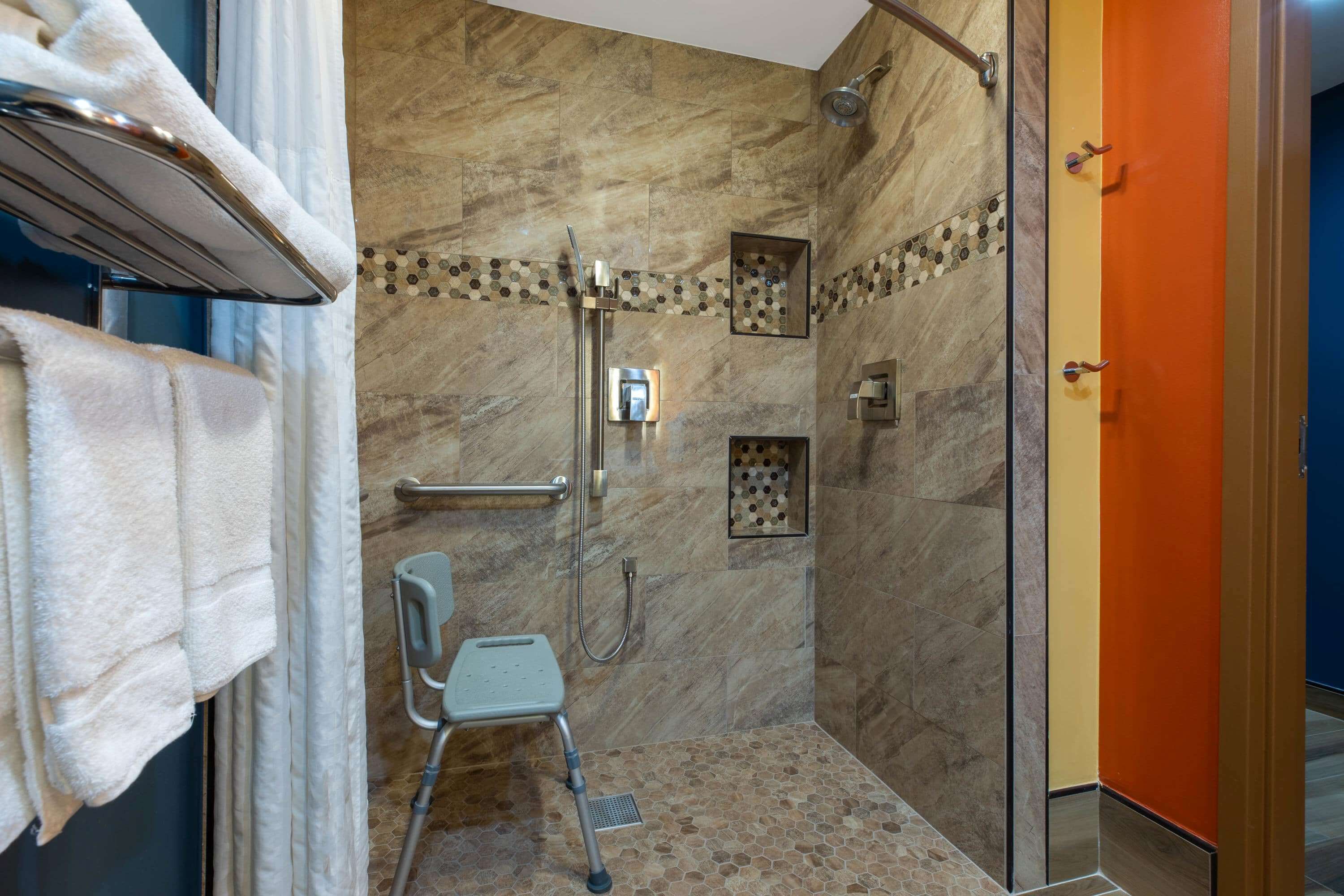Uma área de chuveiro com barras de apoio, chuveiro e um pequeno banquinho, além de piso revestido com azulejos.