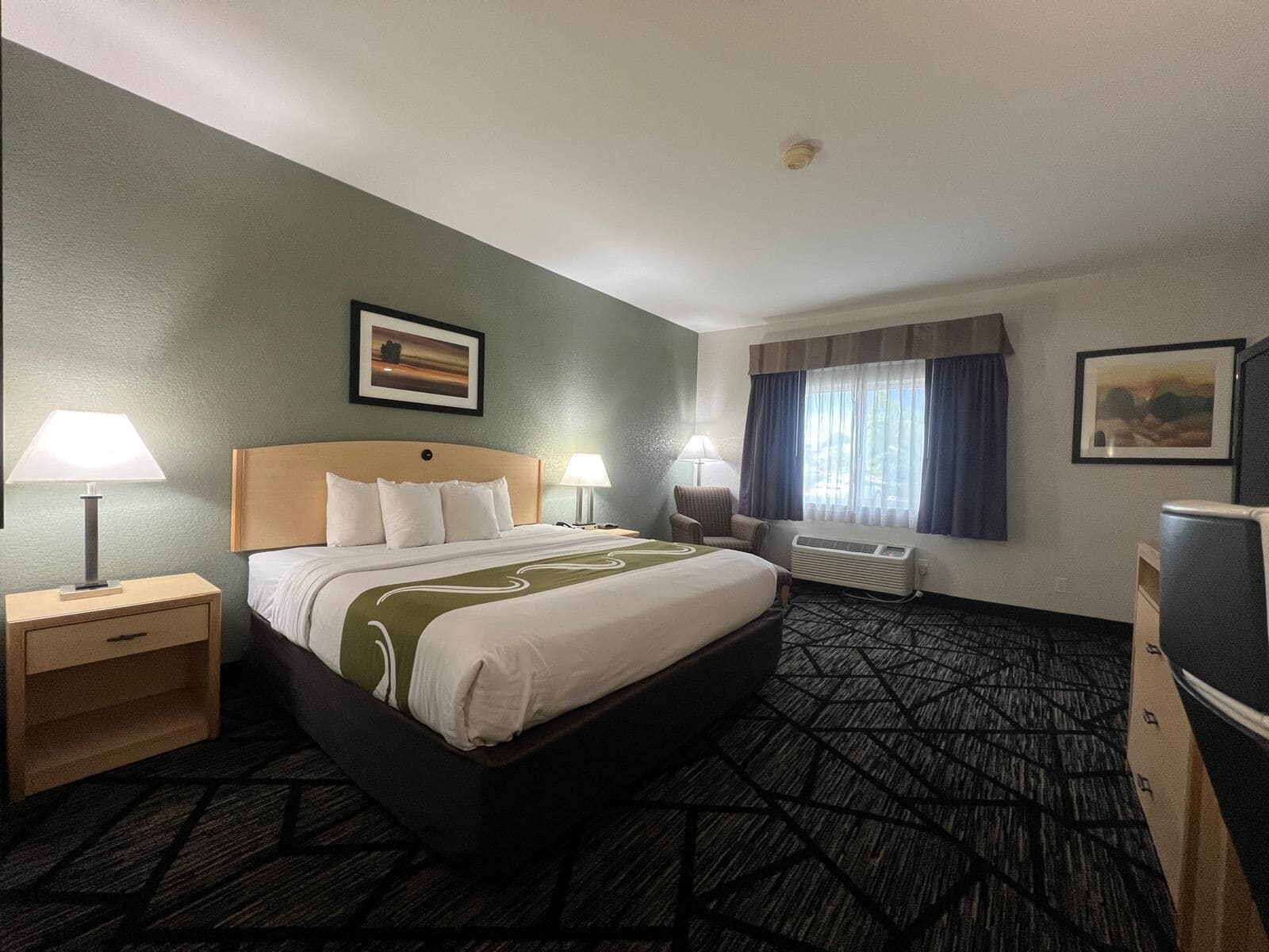 Quarto de hotel com cama, duas mesas de cabeceira, escrivaninha, cadeira, janela com cortinas e televisão presa à parede.