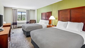 Quarto de hotel com duas camas, uma escrivaninha com computador, uma luminária e vista para o exterior.