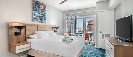 Quarto de hotel com cama, televisão, ventoinha no teto, mesa de cabeceira, cômoda e varanda com vista.