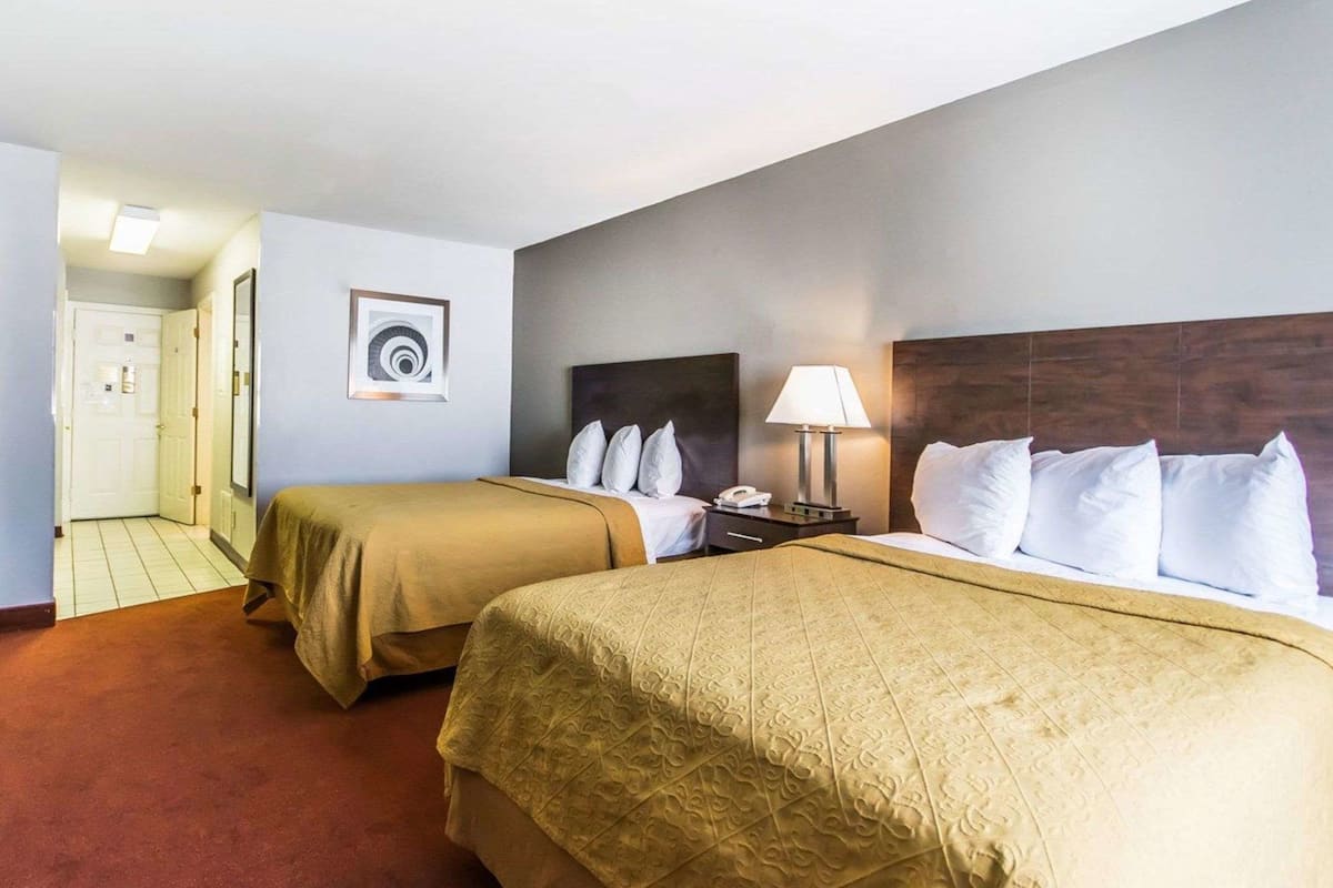 Quarto de hotel com duas camas, uma mesa de cabeceira com abajur, um quadro na parede e uma porta que dá para outro cômodo.
