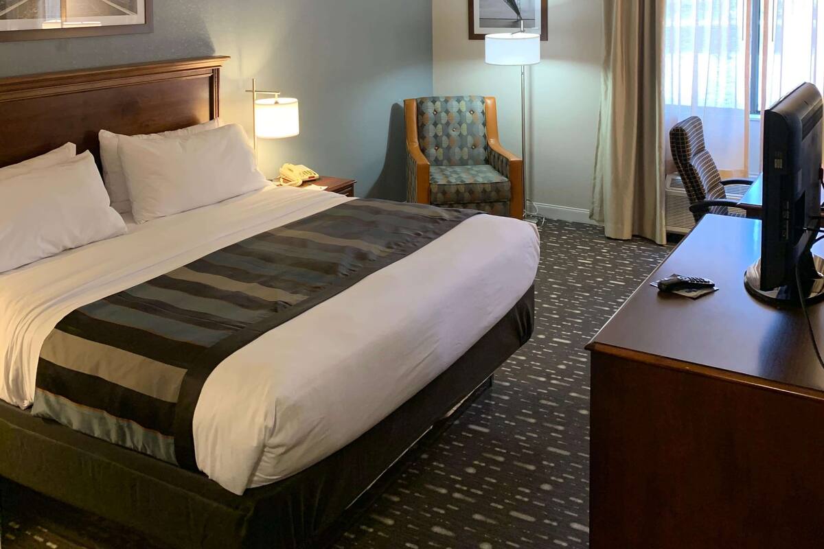Quarto de hotel com uma cama grande, uma mesa com computador, uma cadeira e uma luminária.