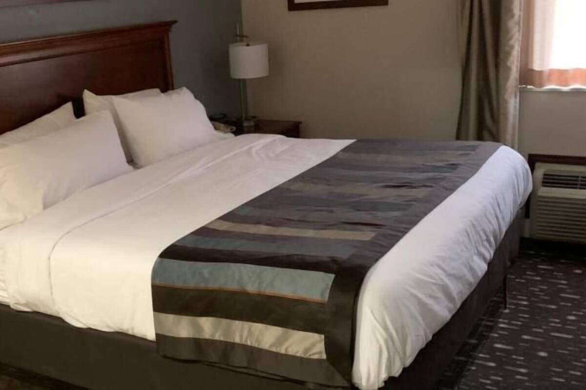 Quarto de hotel com uma cama grande, cabeceira de madeira, mesa de cabeceira com abajur e janela com cortinas.