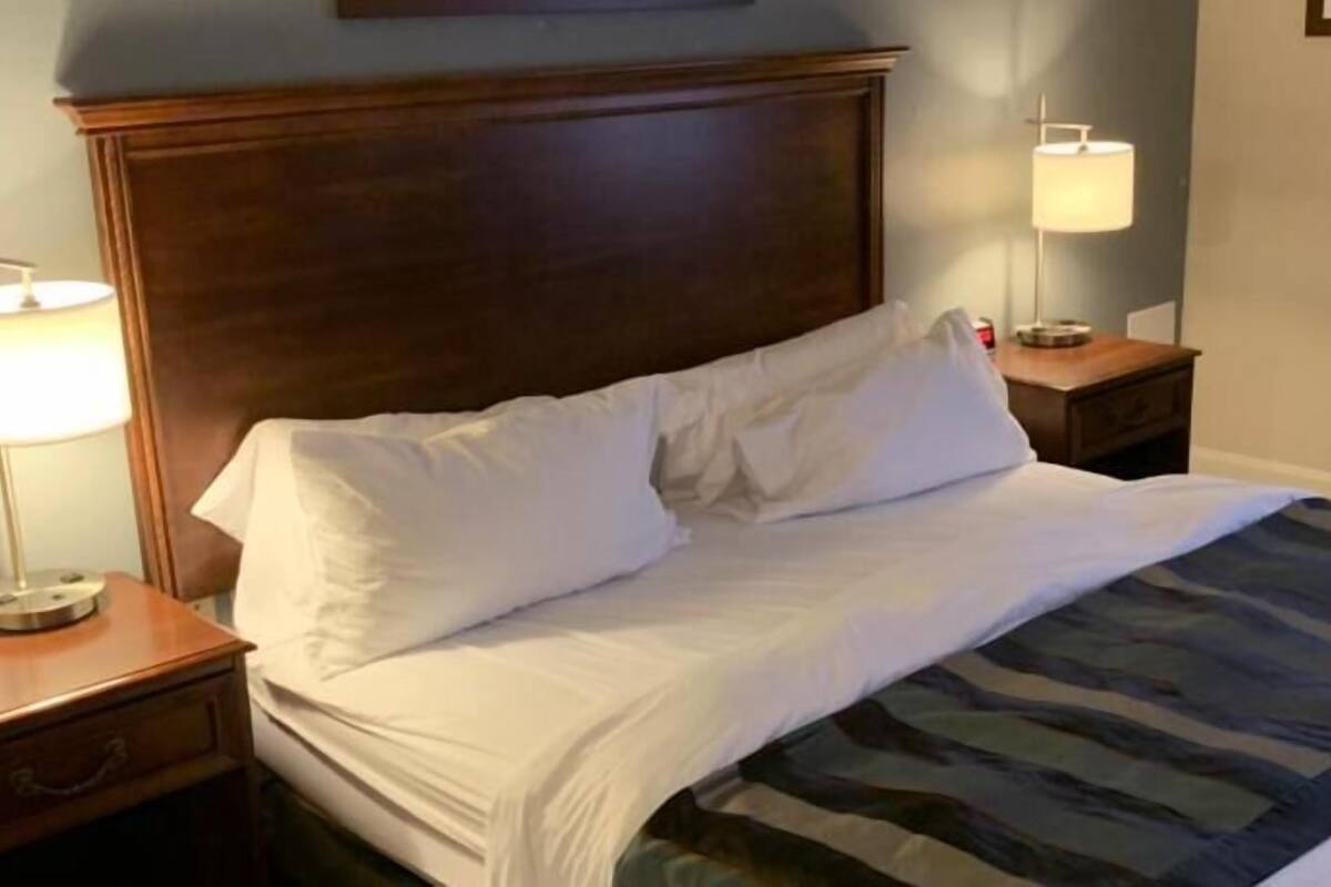 Cama bem arrumada com lençóis brancos, cabeceira de madeira e manta estampada. Há duas mesas de cabeceira com abajures, uma de cada lado da cama.