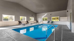 Piscina interior com água azul cristalina, rodeada por espreguiçadeiras e mesas, e grandes janelas que proporcionam luz natural.