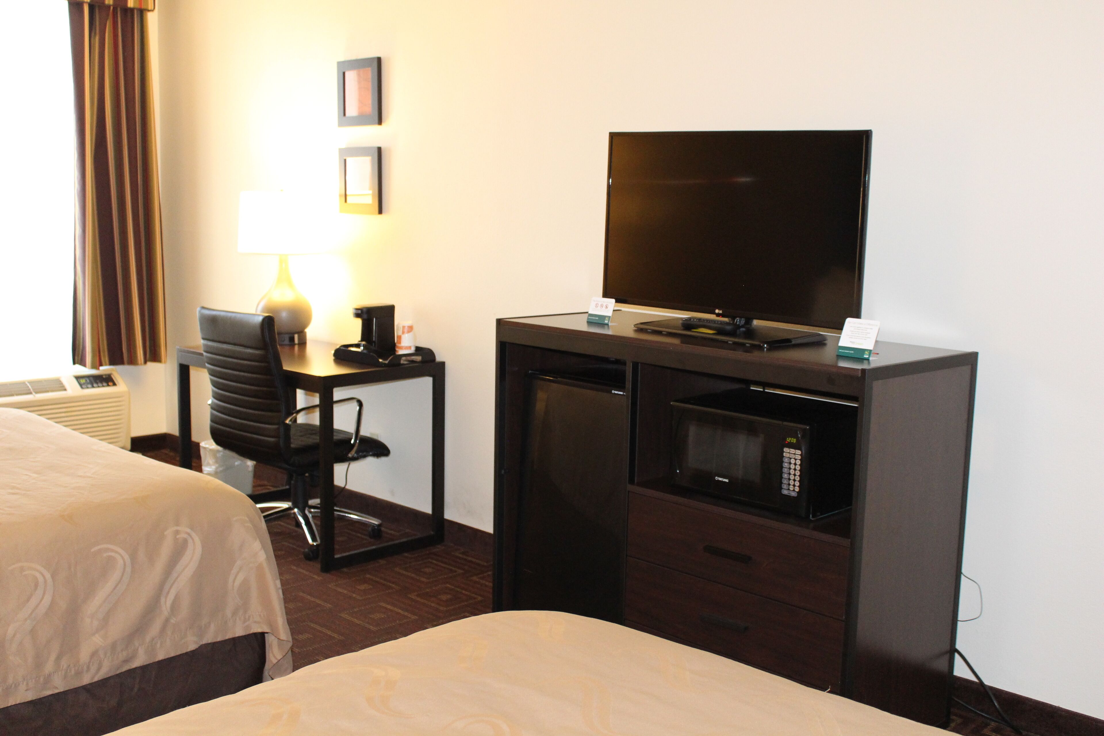 Quarto de hotel com cama, escrivaninha com luminária, televisão, refrigerador e micro-ondas.