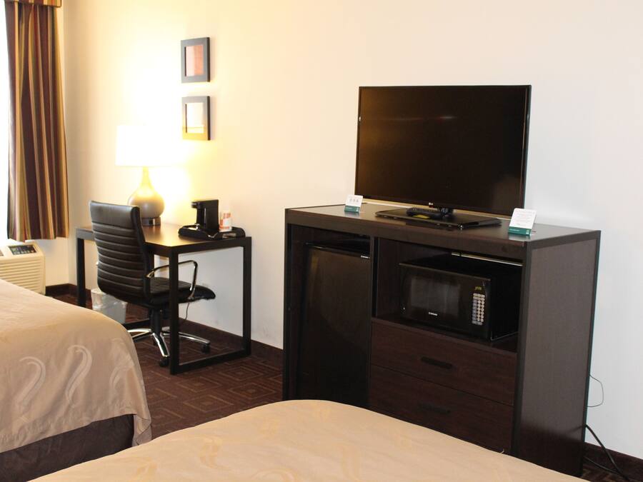 Quarto de hotel com cama, escrivaninha com luminária, televisão, refrigerador e micro-ondas.