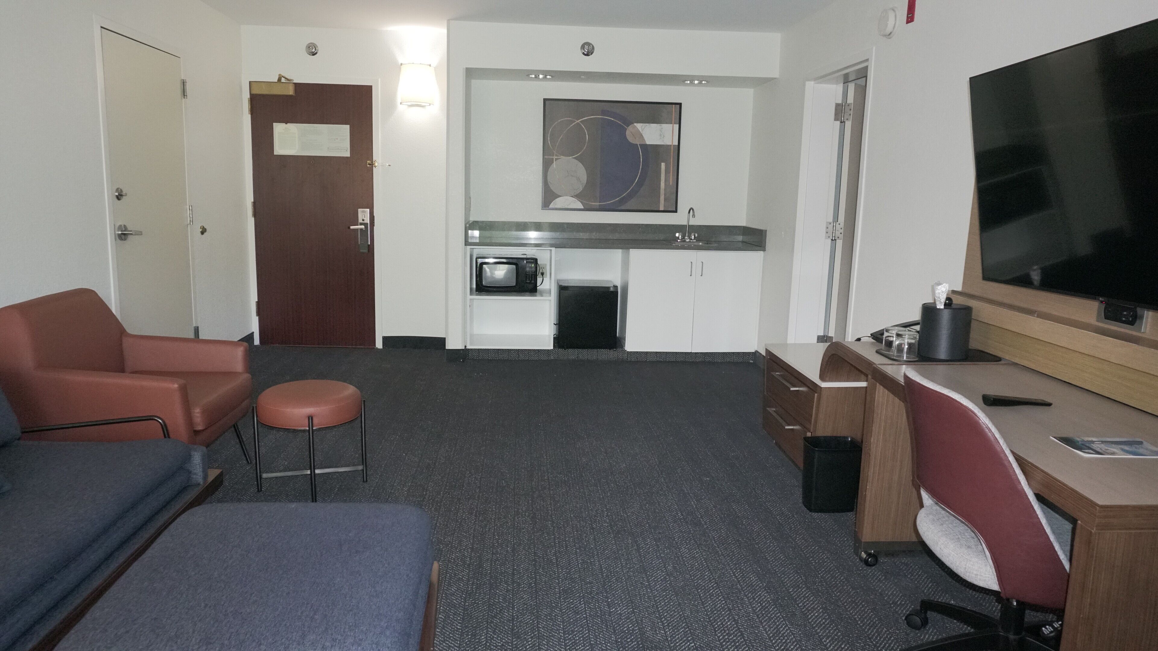 Quarto de hotel pequeno com cozinha compacta, televisão, escrivaninha e área de estar.