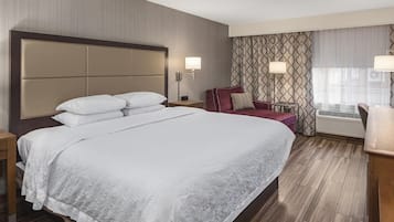 Quarto de hotel com uma cama grande, cabeceira, duas mesas de cabeceira, um sofá vermelho, uma mesinha e uma janela com cortinas.