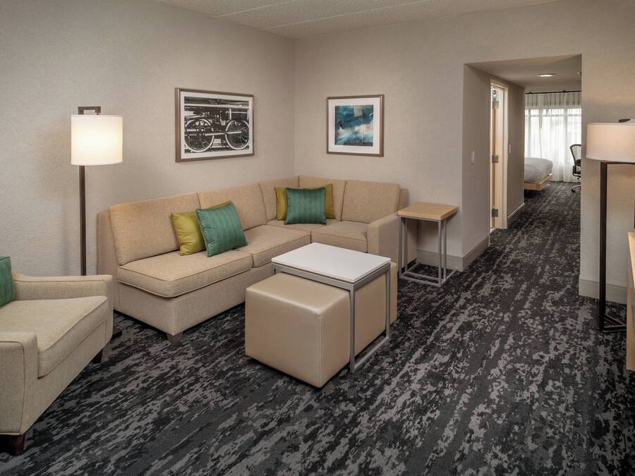 Quarto de hotel com sofá bege, uma mesa pequena, televisão e um quadro na parede.