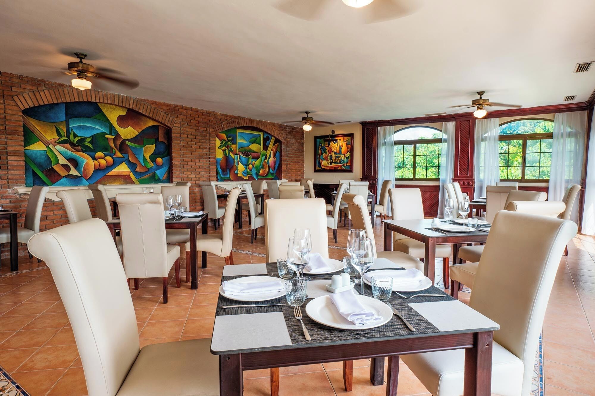 Restaurante com pé-direito alto, paredes de tijolo e pinturas abstratas coloridas. Mesas montadas com toalhas brancas, pratos e copos.