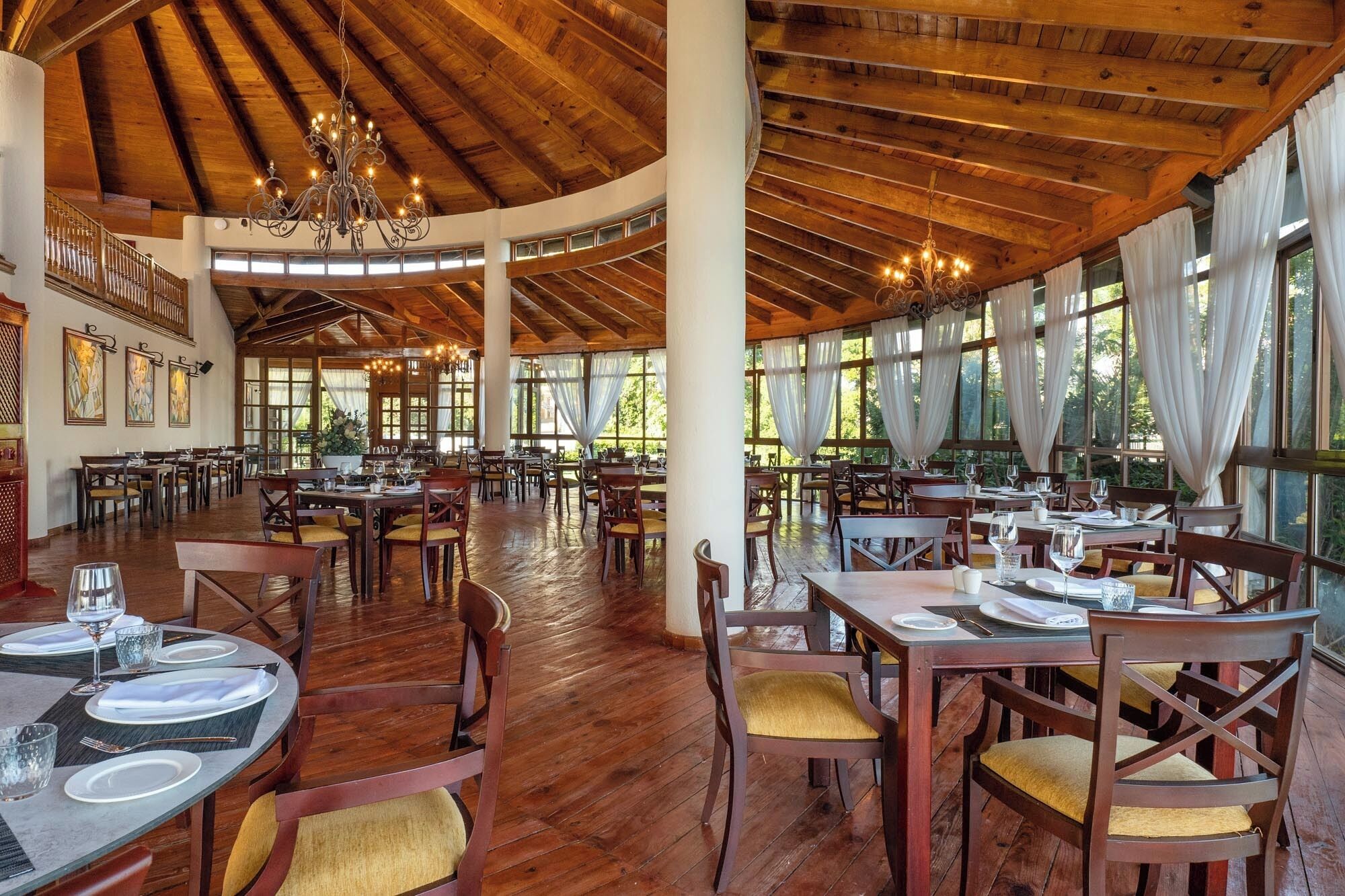 Um restaurante espaçoso com altos tetos de madeira e amplas janelas. As mesas estão dispostas com pratos, copos e talheres.