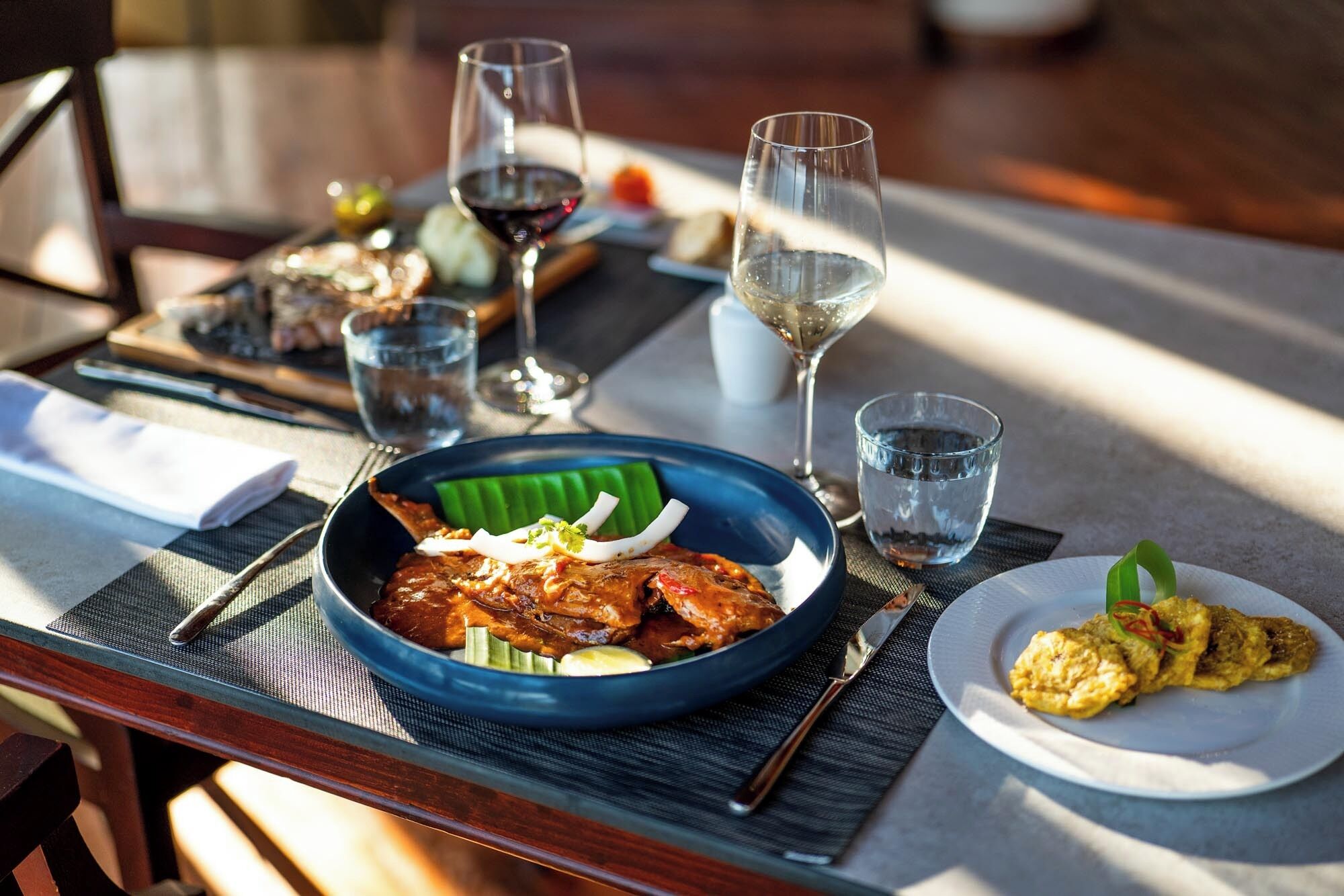 Uma mesa de jantar com uma tigela azul de curry, um prato de comidas fritas e copos de água e vinho.
