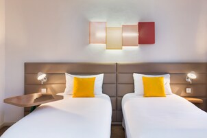 Quarto de hotel com duas camas, cada uma com roupa de cama branca e um travesseiro amarelo. Há luminárias fixadas na parede acima das camas e uma mesa de cabeceira de madeira.