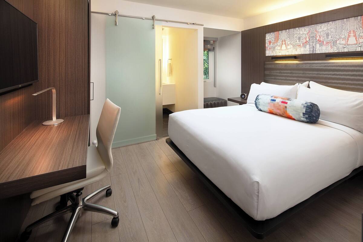 Quarto de hotel com cama, secretária, cadeira e televisão.