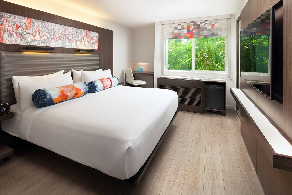 Quarto de hotel moderno com uma cama grande, uma escrivaninha, uma cadeira, uma TV e uma janela com vista para o verde.